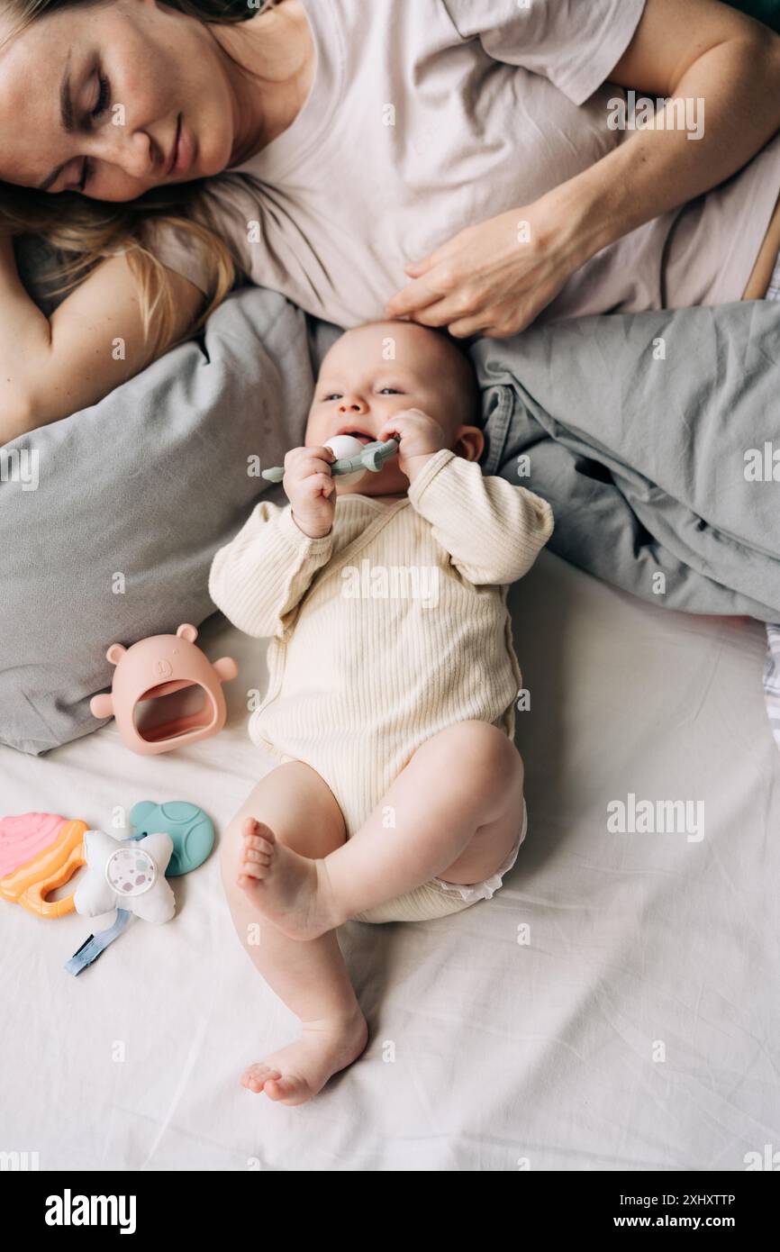 Cute baby plays and gnaws a teething ring toy while mom is lying nearby ...