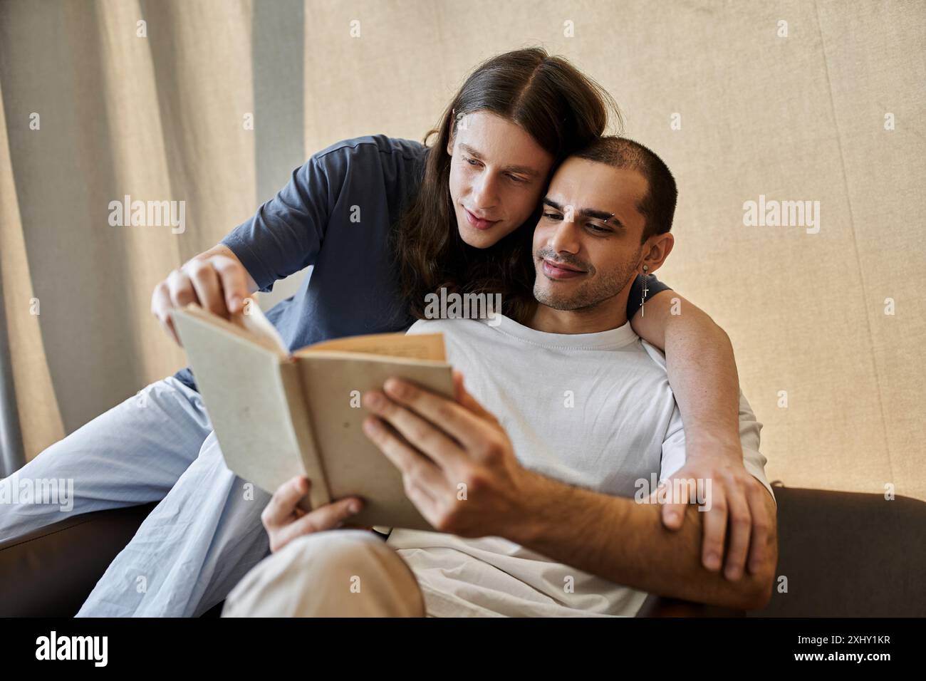 Two young men cuddle on a couch, one reading a book while the other ...