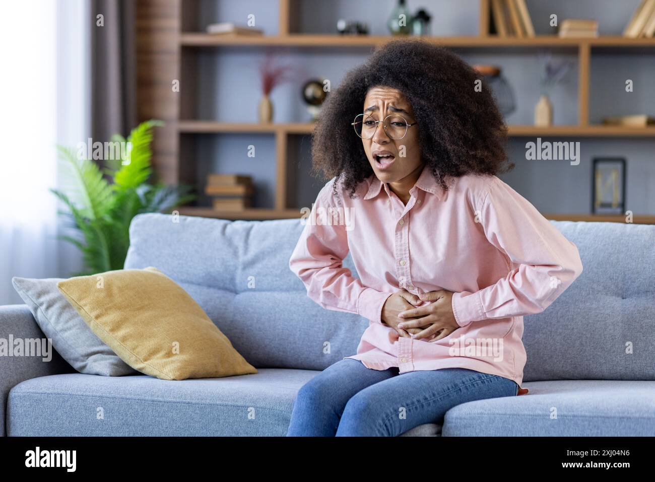Young woman sitting on couch holding stomach in pain. Suffering from ...