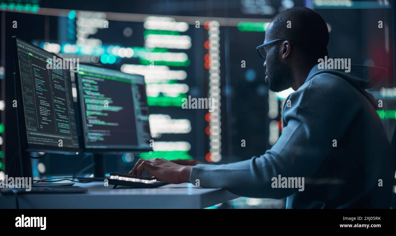 Black Male Programmer Working in Monitoring Room, Surrounded by Big Screens Displaying Lines of ...
