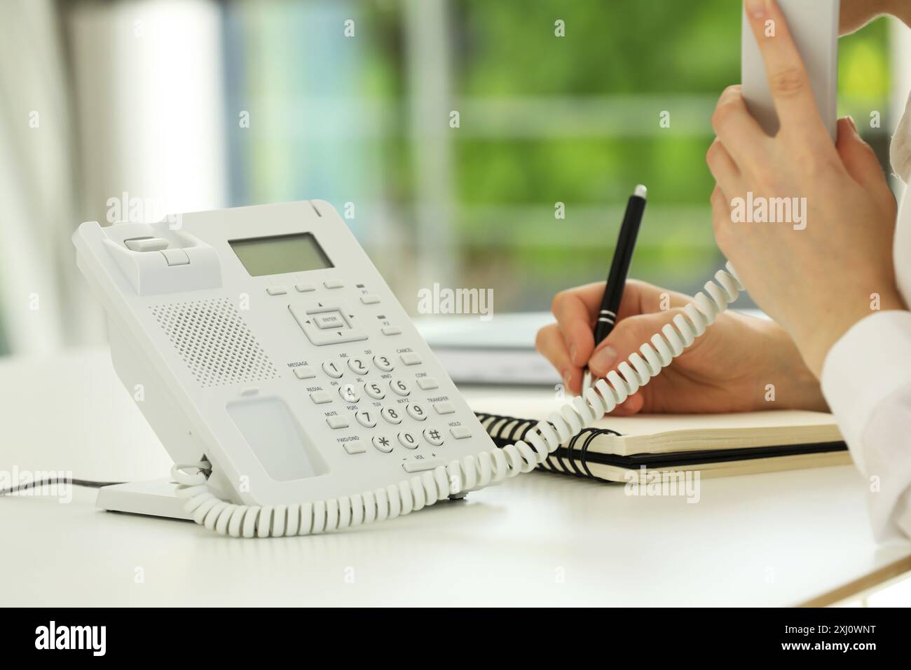 Assistant with telephone handset writing at white table against blurred ...