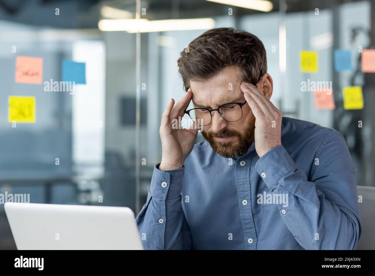 A tired and overworked young man is sitting at the desk in the office, grimacing in pain and ...