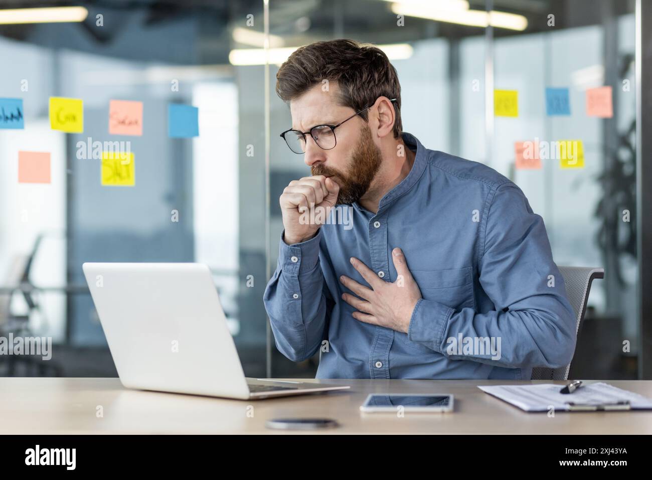 A young male office worker sits at a desk at work and coughs, covers ...