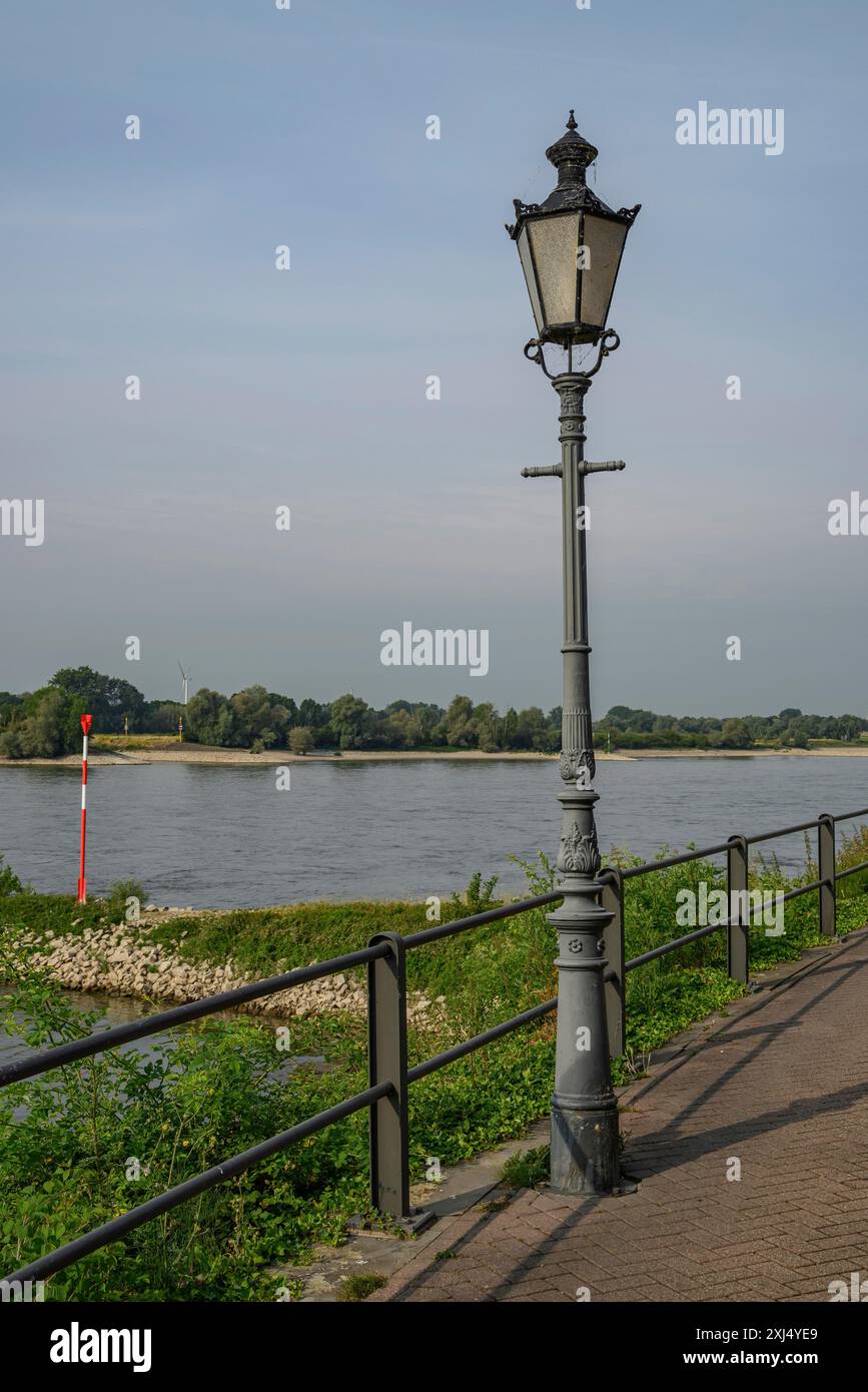 An old street lamp stands on the bank of a river, along a paved ...