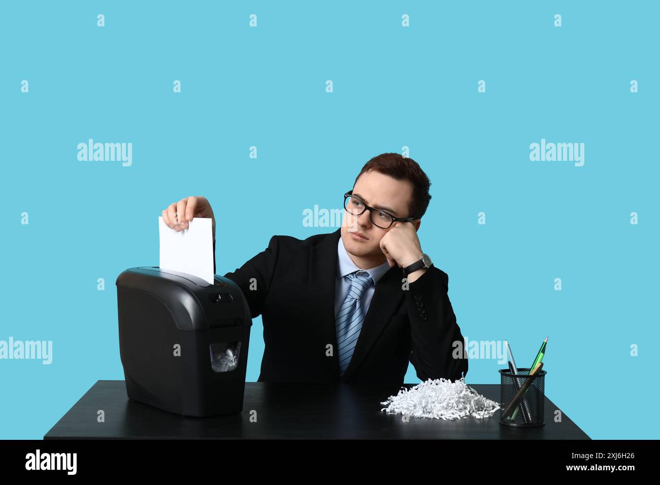 Sad young man destroying paper sheet in shredder on blue background ...