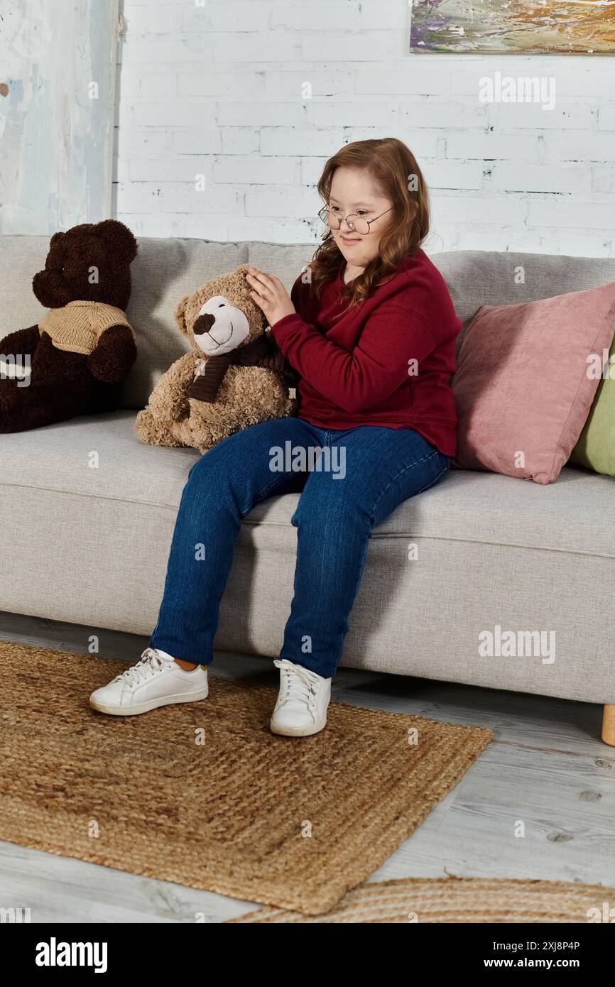 A little girl with Down syndrome sits on a couch with two teddy bears ...