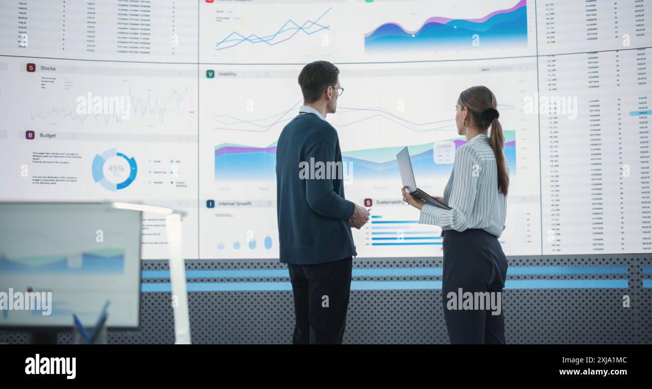 Back View Following Shot Of Male And Female Analysts Discussing Data On Big Digital Screen In ...
