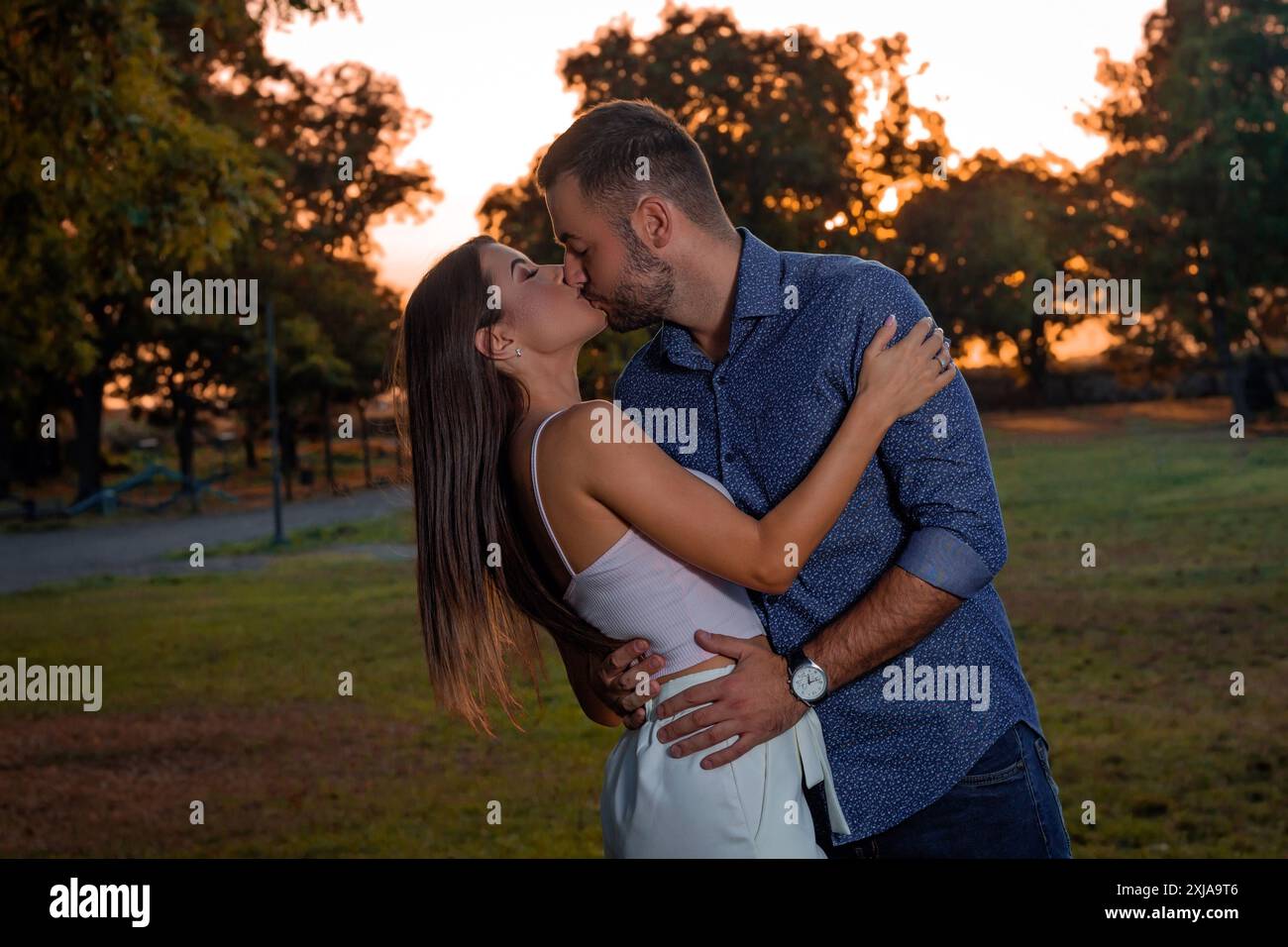 A man and woman embrace and kiss while standing in a park as the sun ...