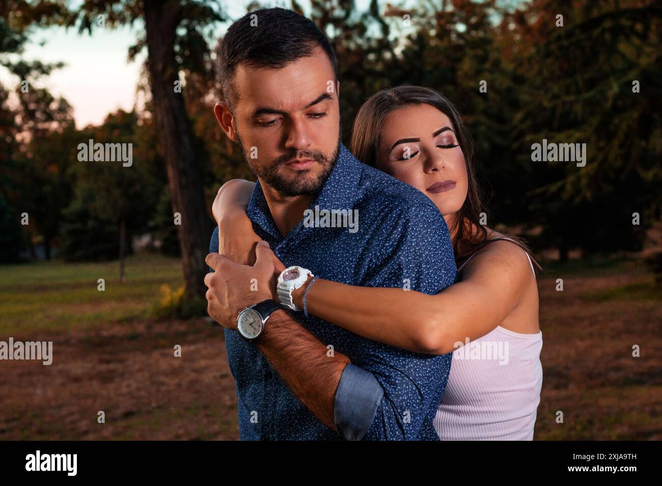 A woman hugs a man from behind as they stand in a park, the golden ...