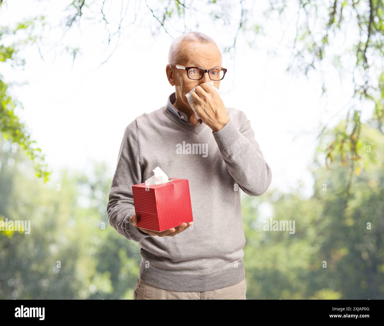 Elderly man blowing nose and holding a box of paper tissues outdoors in ...