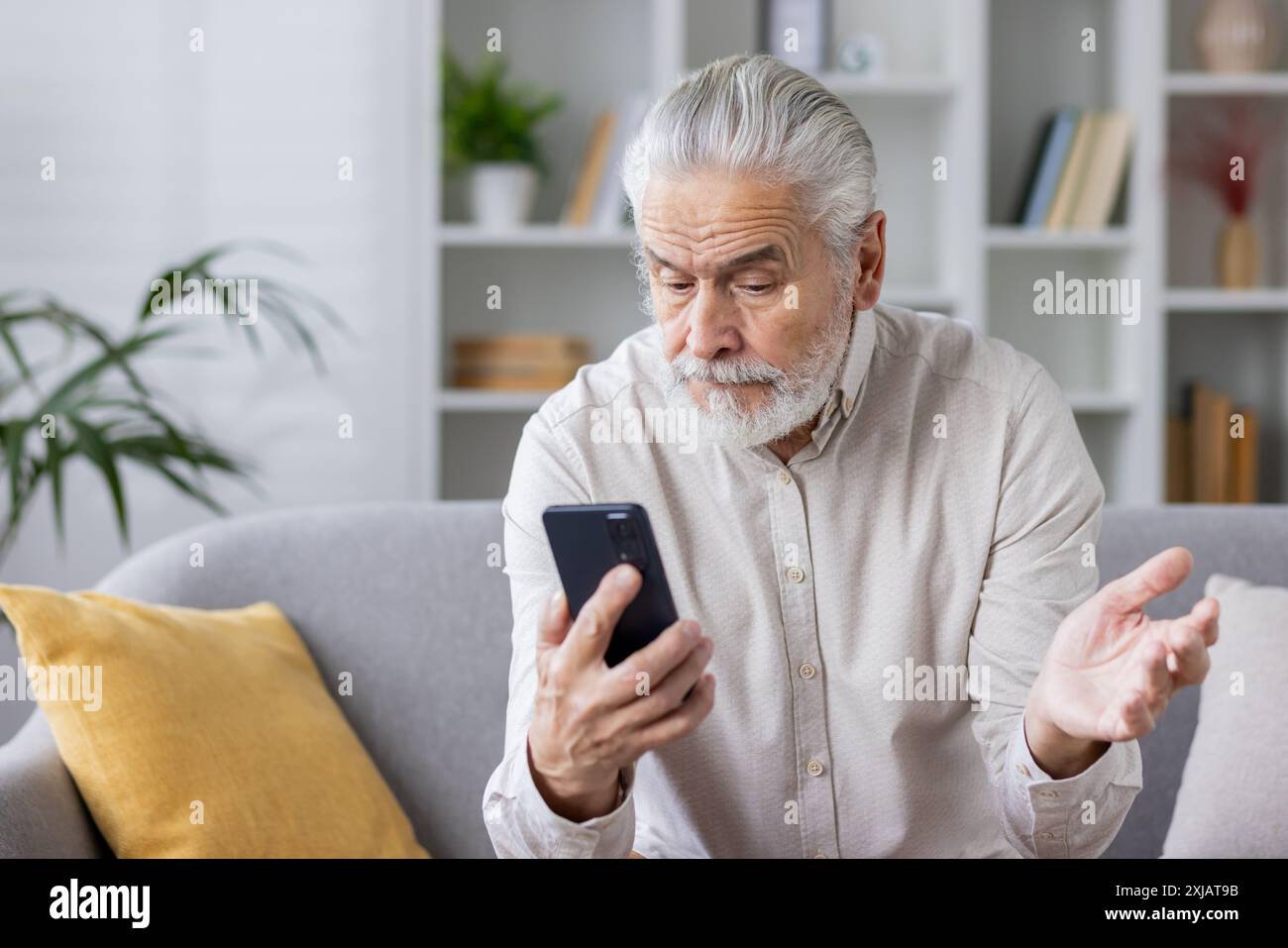 Elderly man sitting on couch using mobile phone, looking confused and ...