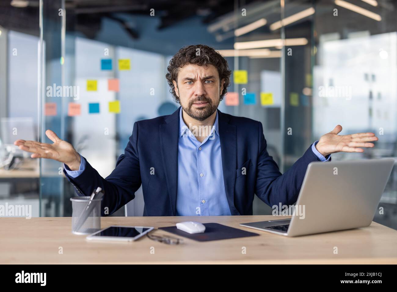 Confused businessman sitting at desk in modern office, shrugging with ...