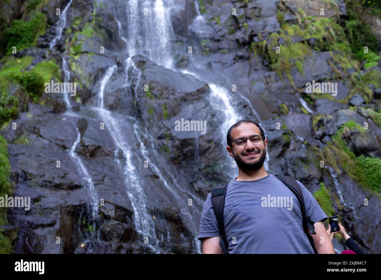 A smiling man stands near a cascading waterfall on wet, dark rocks ...