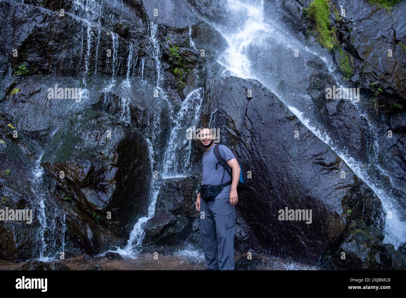 A smiling man stands near a cascading waterfall on wet, dark rocks ...