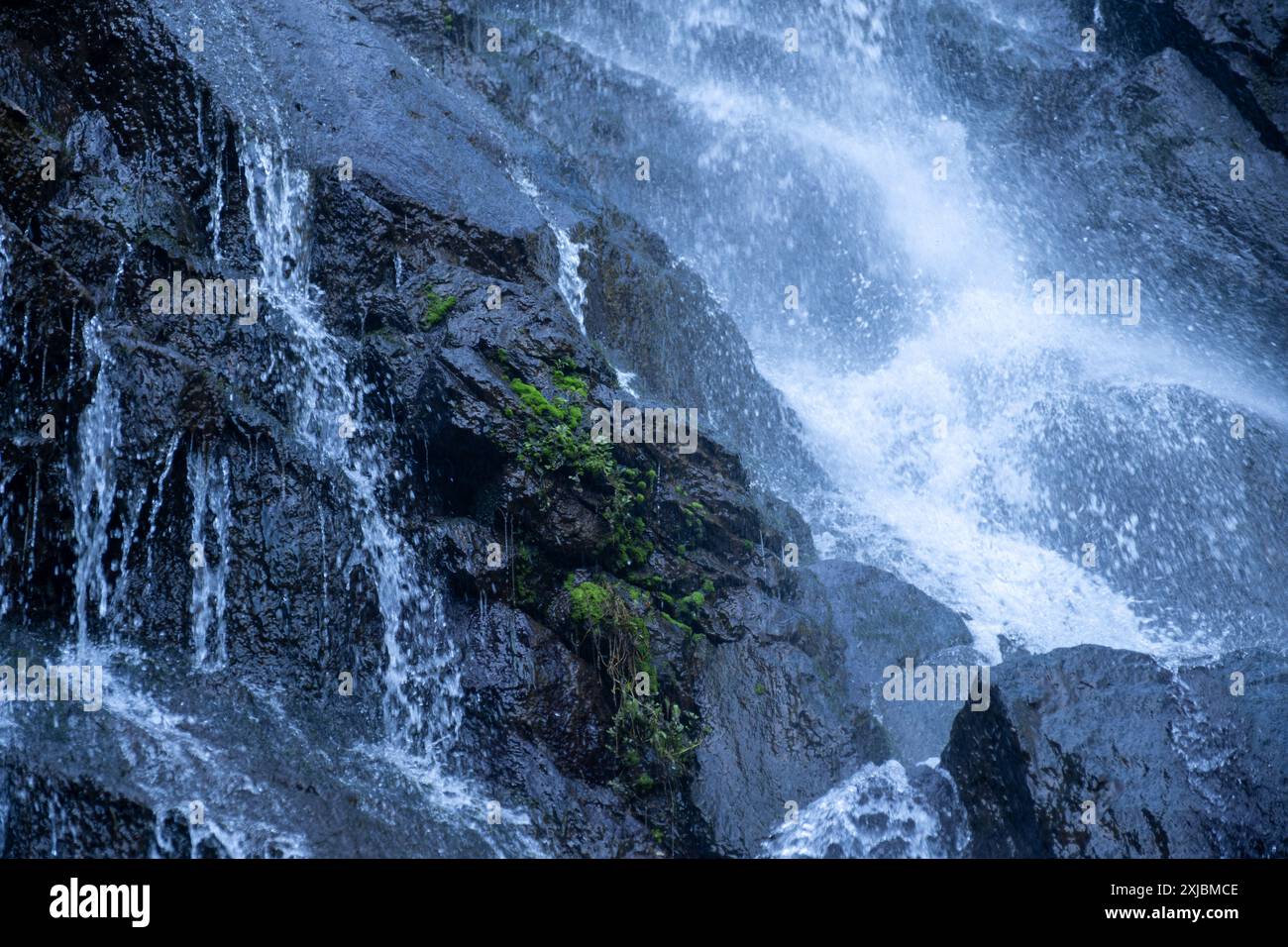 A smiling man stands near a cascading waterfall on wet, dark rocks ...