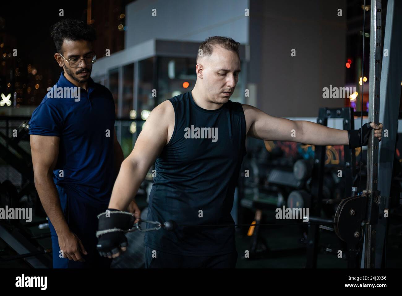 A man works out on a cable machine while his trainer observes. The gym ...