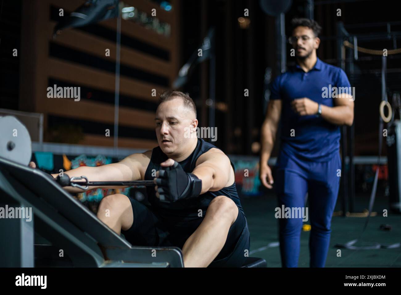 A man works out on a rowing machine at a gym. Another man stands behind ...