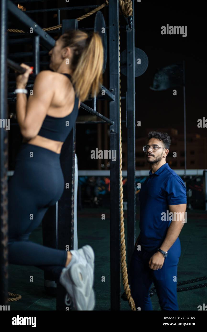 A woman in black workout clothes does pull-ups on a bar while a trainer ...