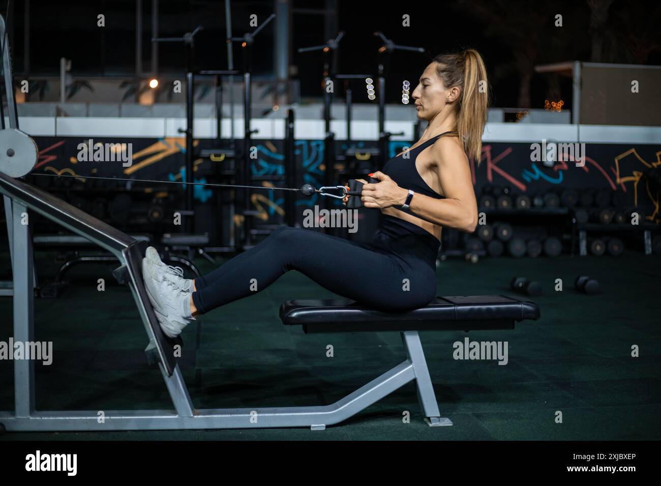 A woman is working out in a gym. She's seated on a cable machine and ...