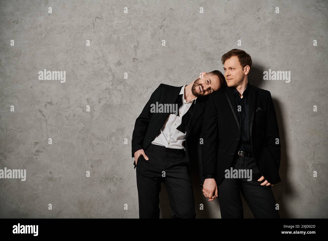 Two men in elegant black suits stand against a grey wall, showing their ...