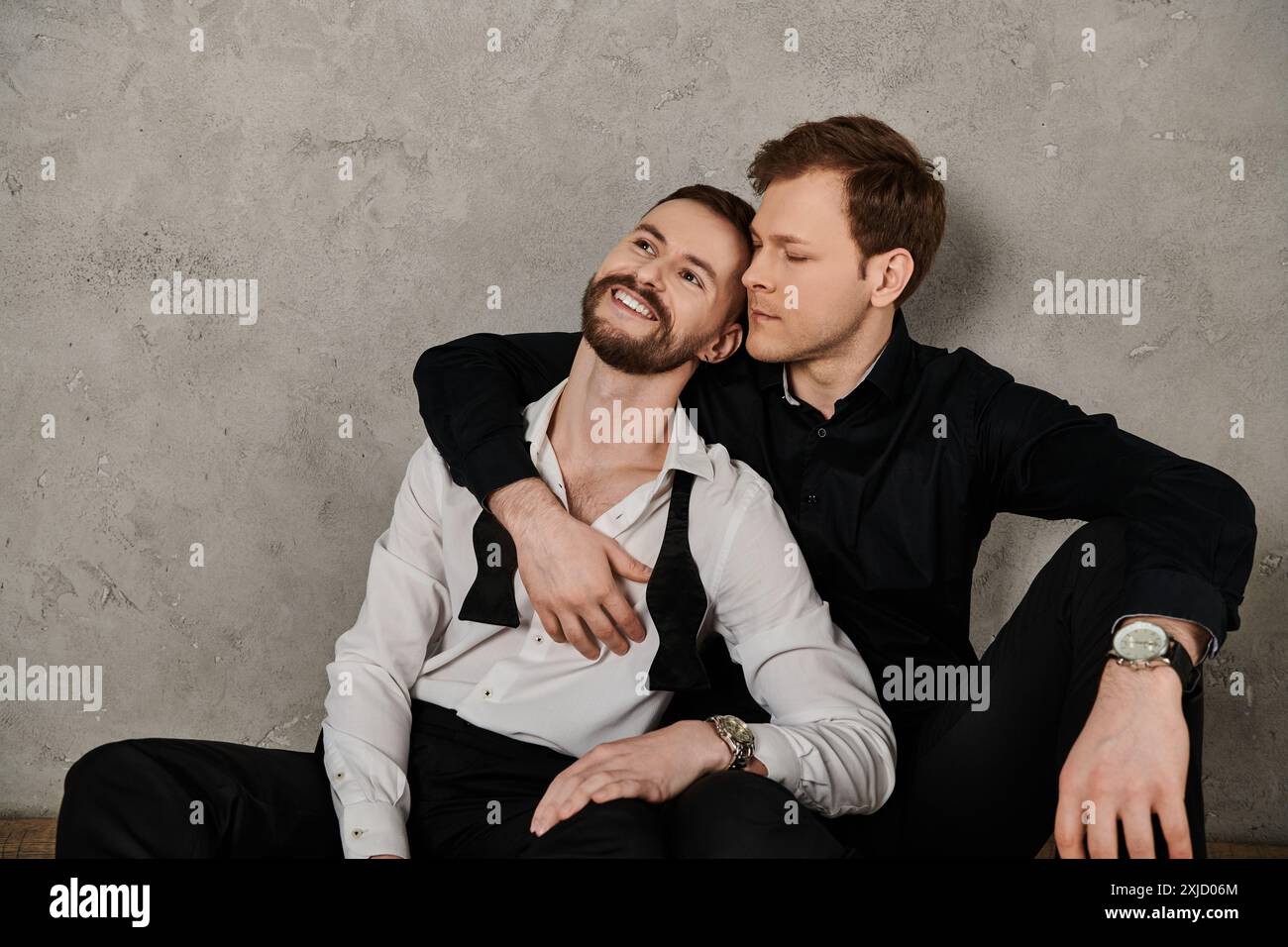 Two men in elegant suits share a tender moment against a concrete wall ...