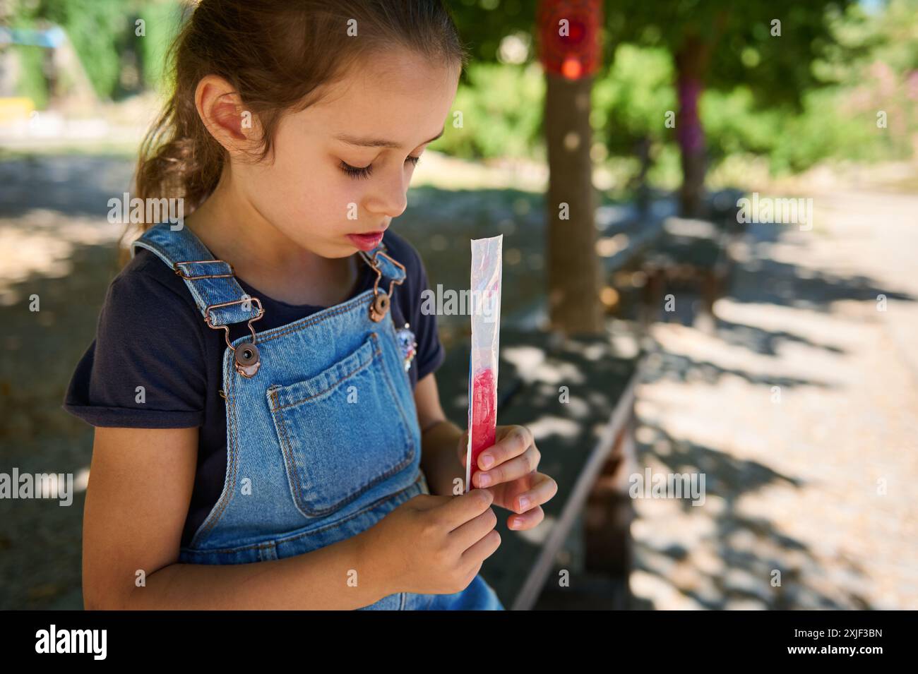 A young girl in denim overalls enjoying a cold popsicle on a warm ...