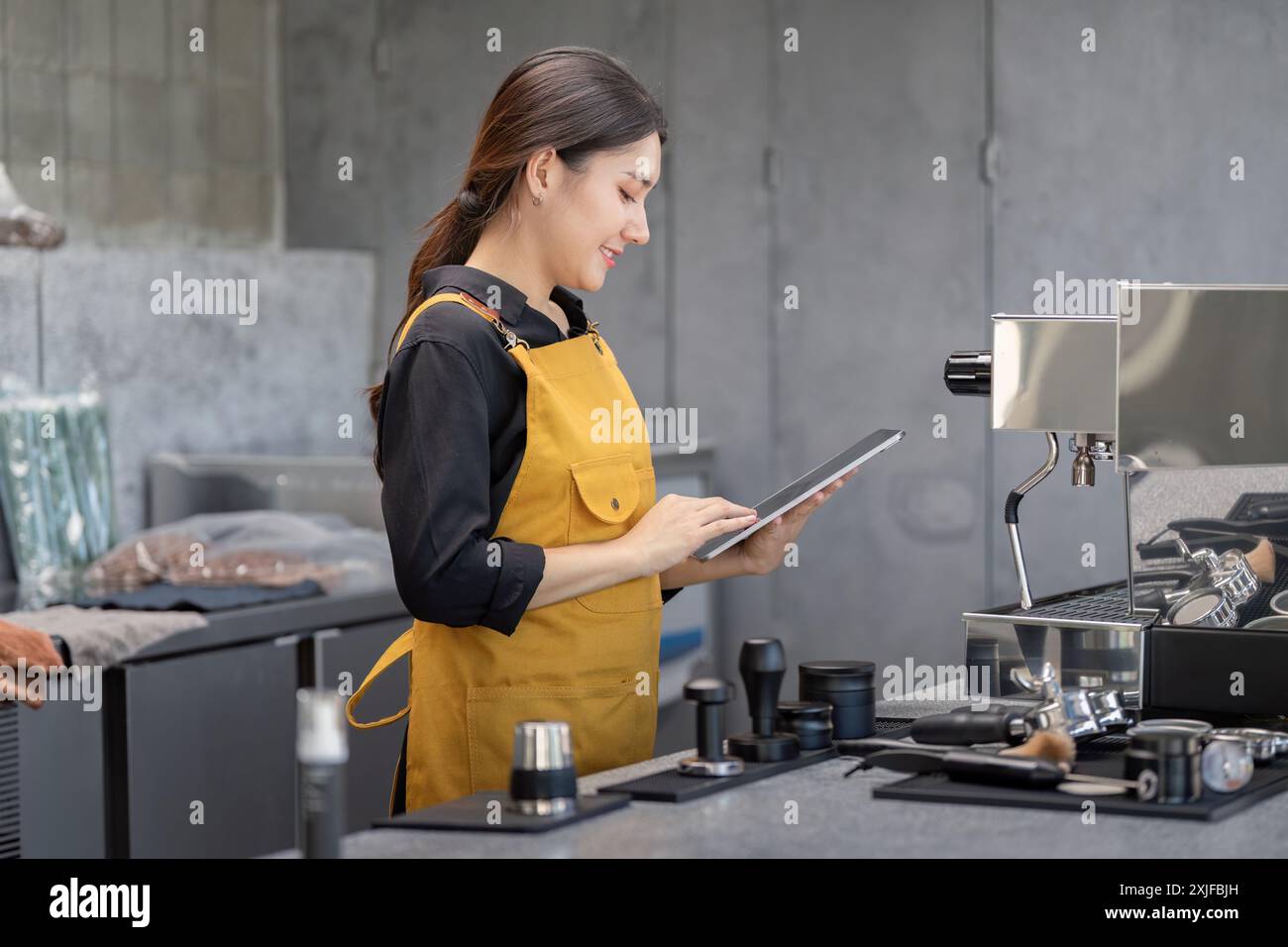 Young Female Barista in Yellow Apron Using Tablet in Modern Coffee Shop with Professional ...
