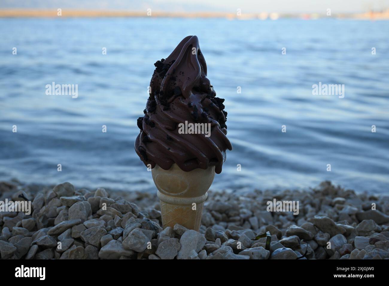 Chocolate ice cream cone melting on a pebble beach with blue water in ...