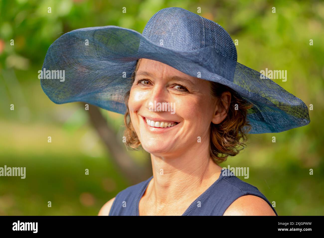 An adult woman with wavy brown hair smiles while wearing a wide-brimmed ...