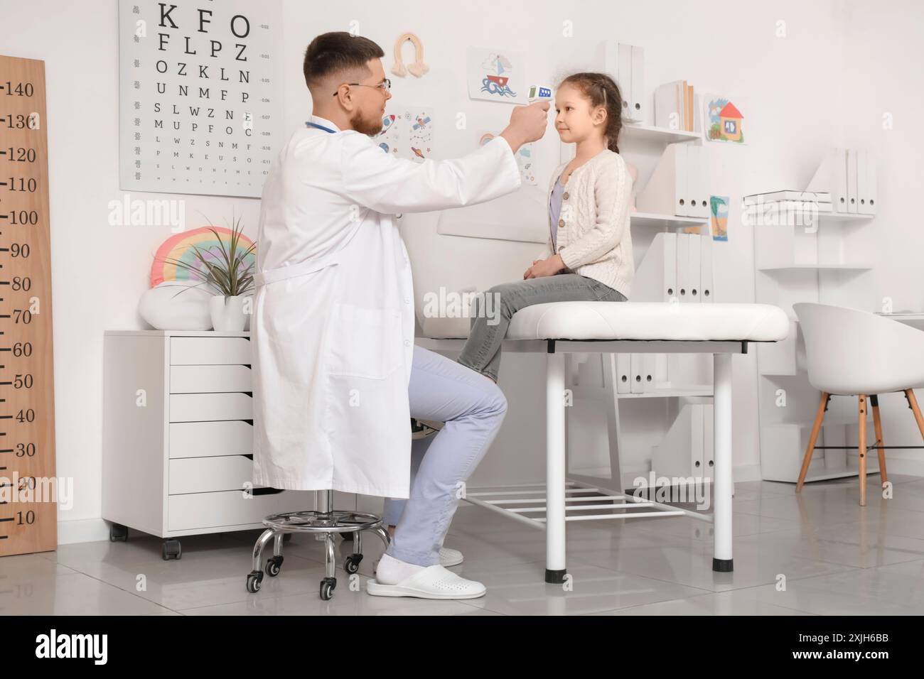 Male pediatrician measuring little girl's temperature on couch in ...