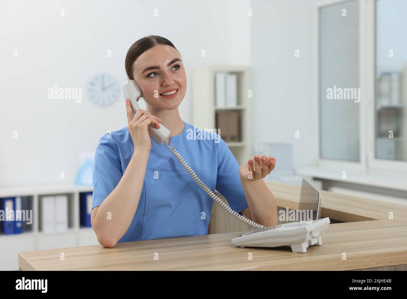 Professional receptionist talking on phone at wooden desk in hospital ...