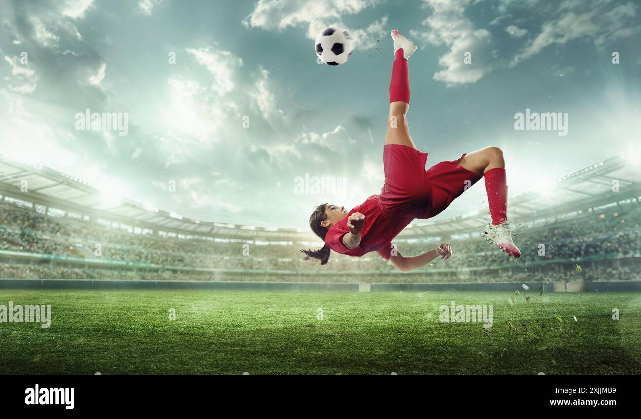 Female soccer player in red performs bicycle kick on stadium field. Stadium with spectators ...