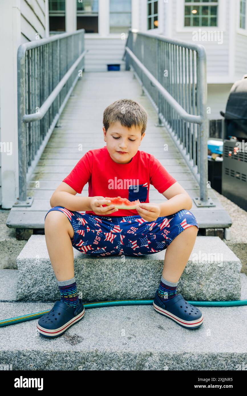 Boy in a red shirt and blue shorts eating watermelon on porch steps ...