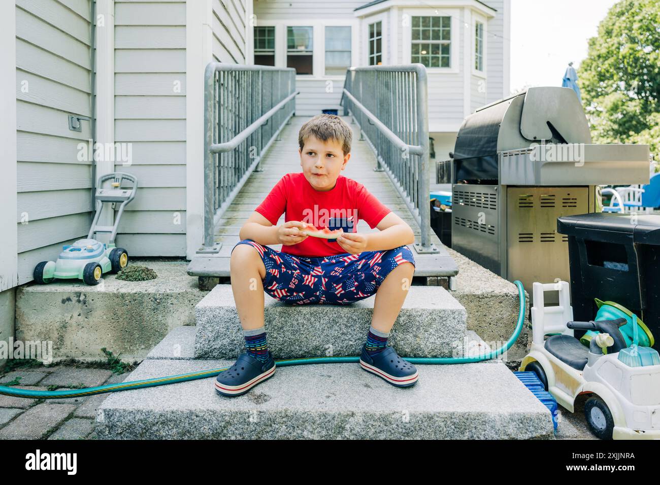 Boy in red shirt and blue shorts eating watermelon with toys around ...