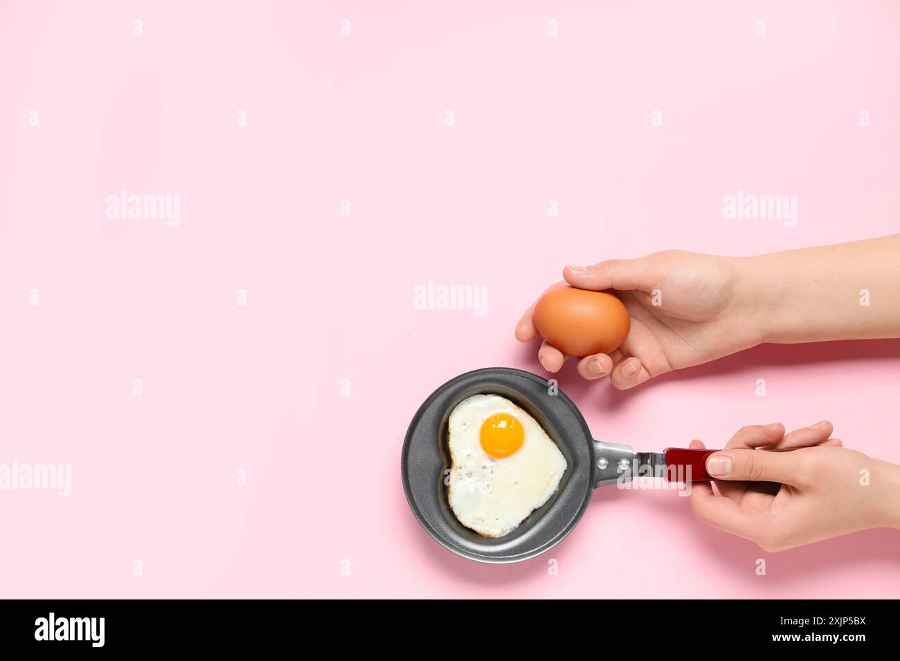 Female hands holding pan with heart-shaped fried egg and egg on pink background Stock Photo - Alamy