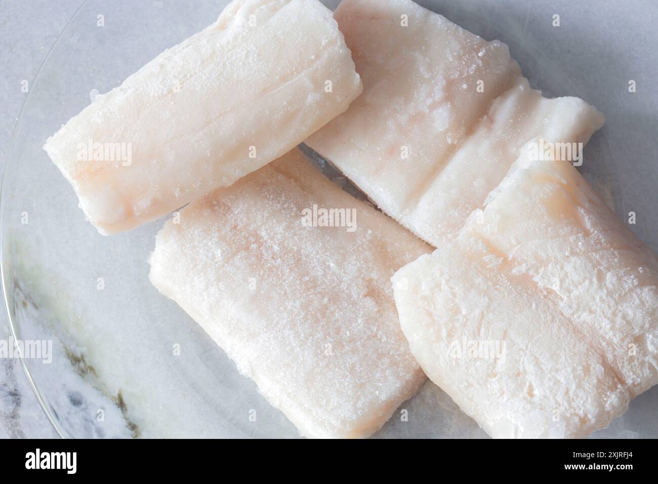 Frozen haddock fillets defrosting on a glass plate. On a light grey ...