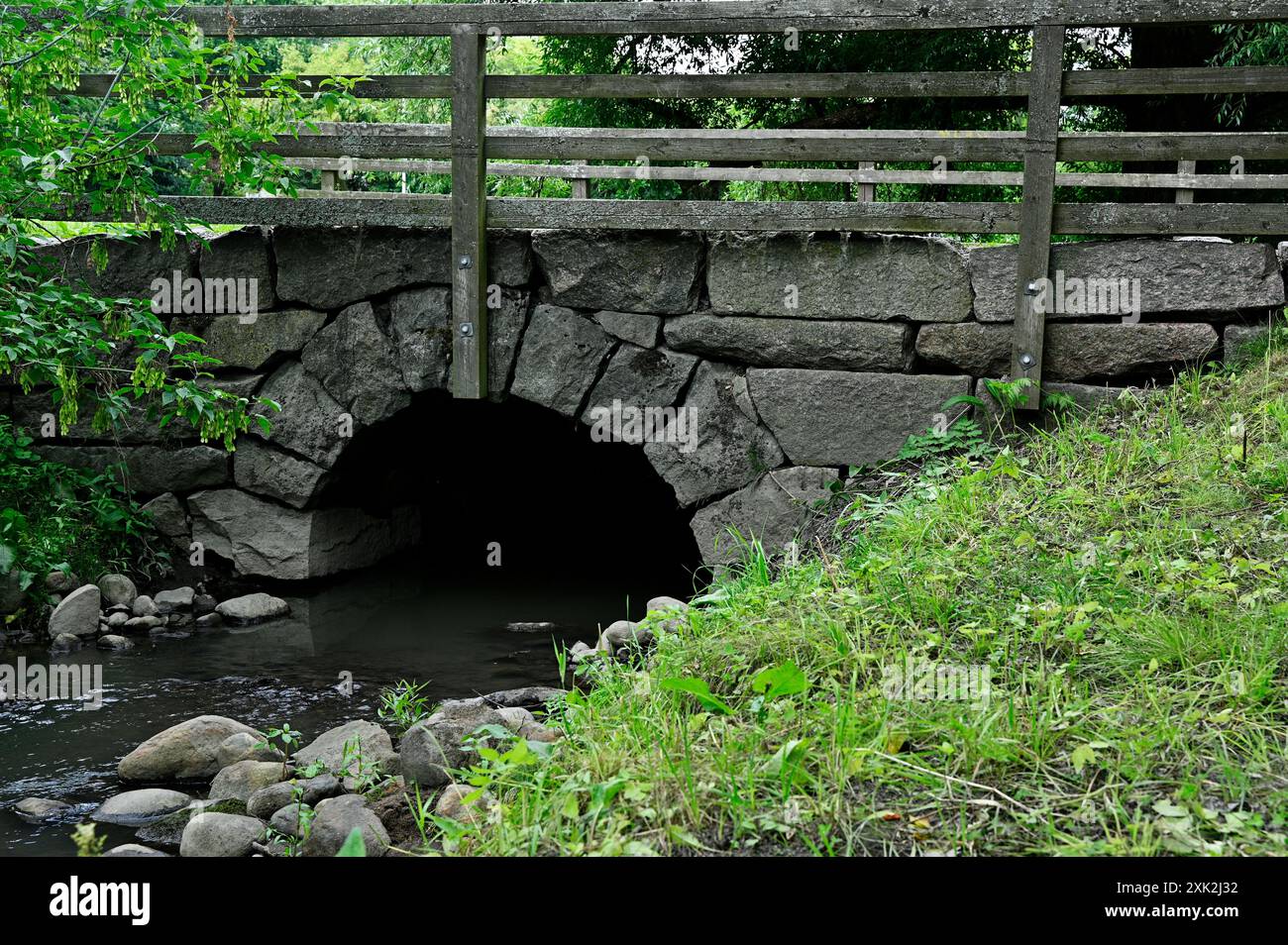 A rustic stone bridge with a wooden railing spans a small creek ...