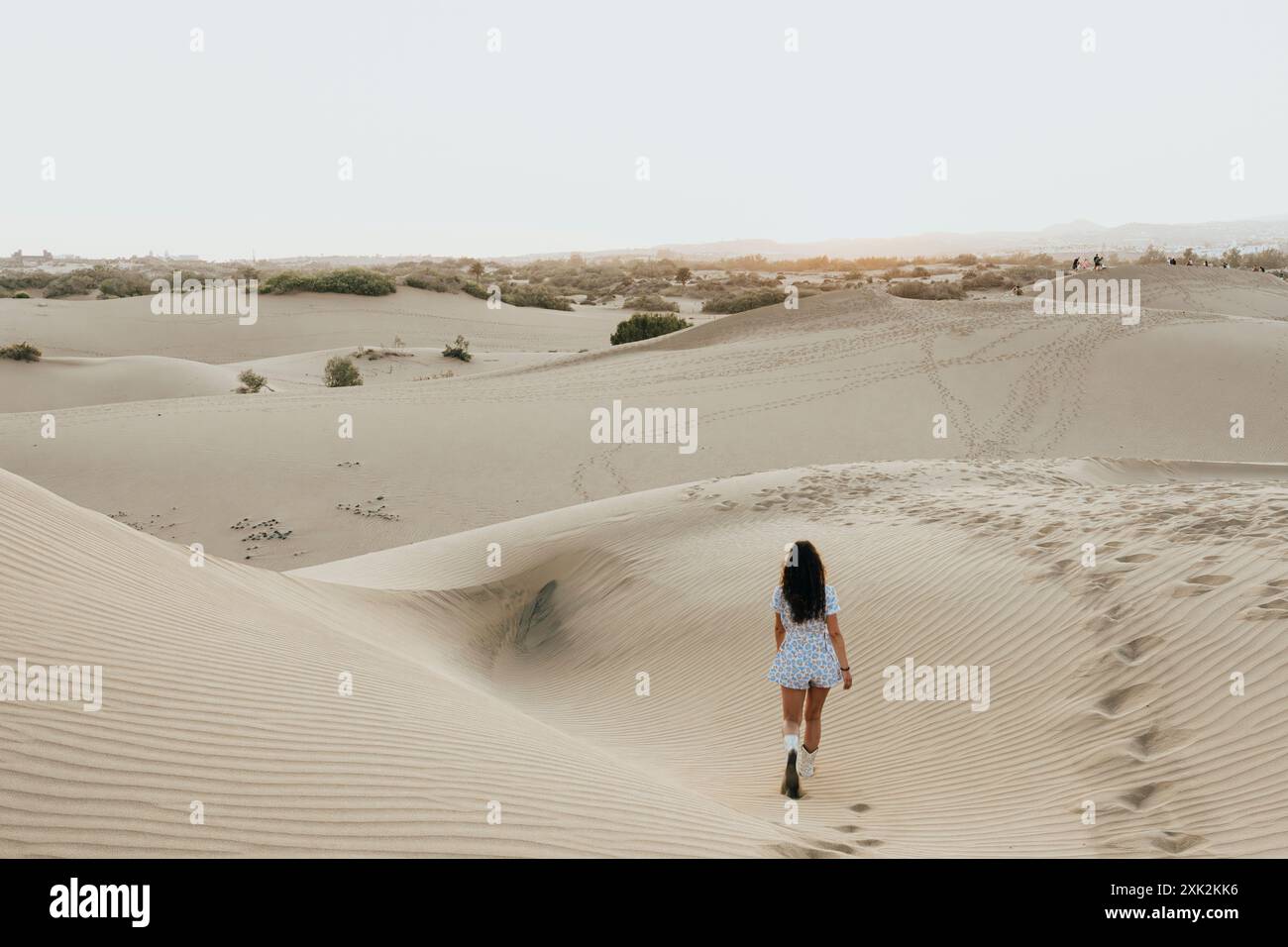 A young woman walks through expansive sand dunes under a soft sunset ...