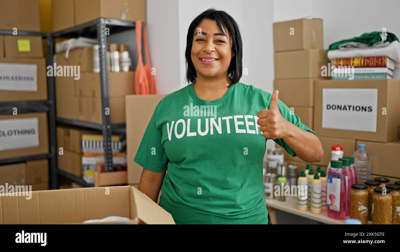 A cheerful hispanic woman volunteer giving a thumbs-up in a warehouse ...