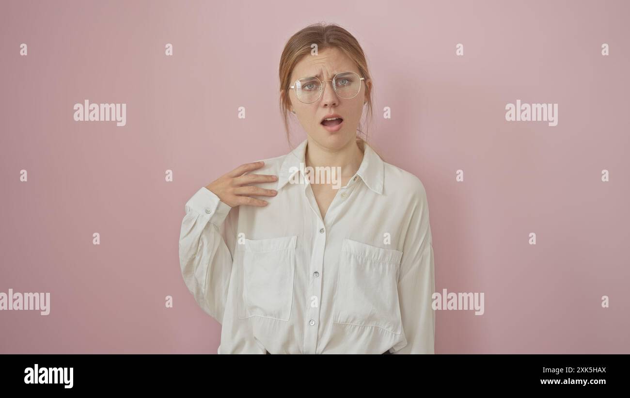 A perplexed young woman with glasses stands against a plain pink ...