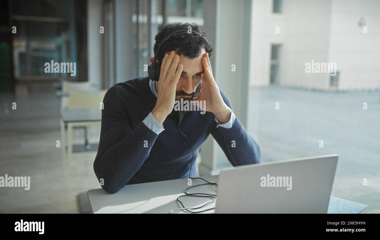 A stressed man with headphones faces a laptop in a modern office ...