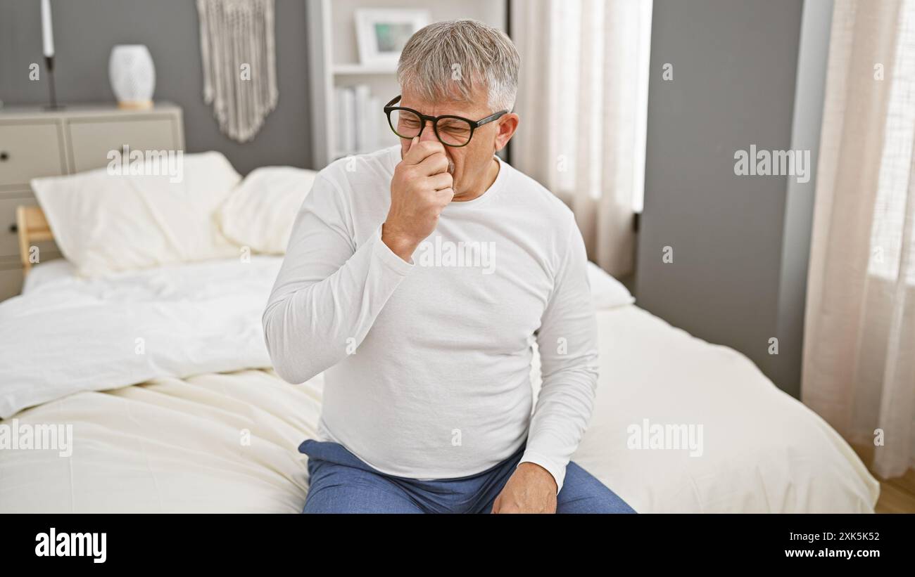 A senior man pinches his nose due to bad odor while sitting on a bed in ...