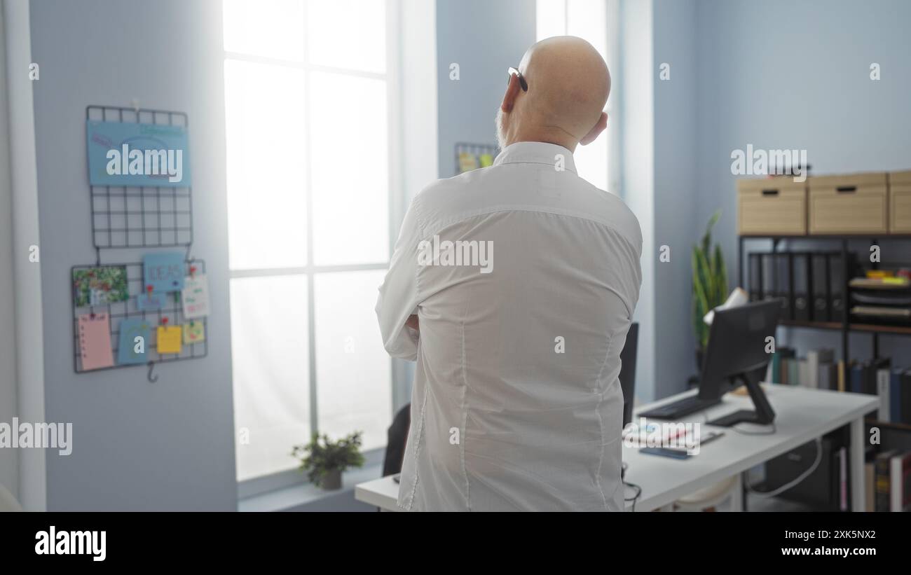 Middle-aged man in an office stands facing a brightly lit window ...