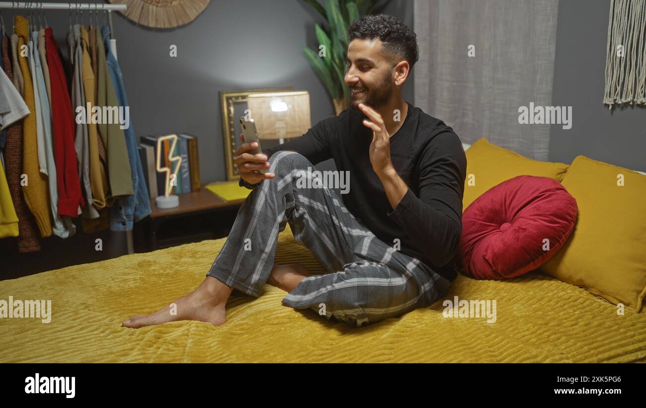 A handsome, young, hispanic man relaxes on a bed in a cozy bedroom, engaging in a video call on ...