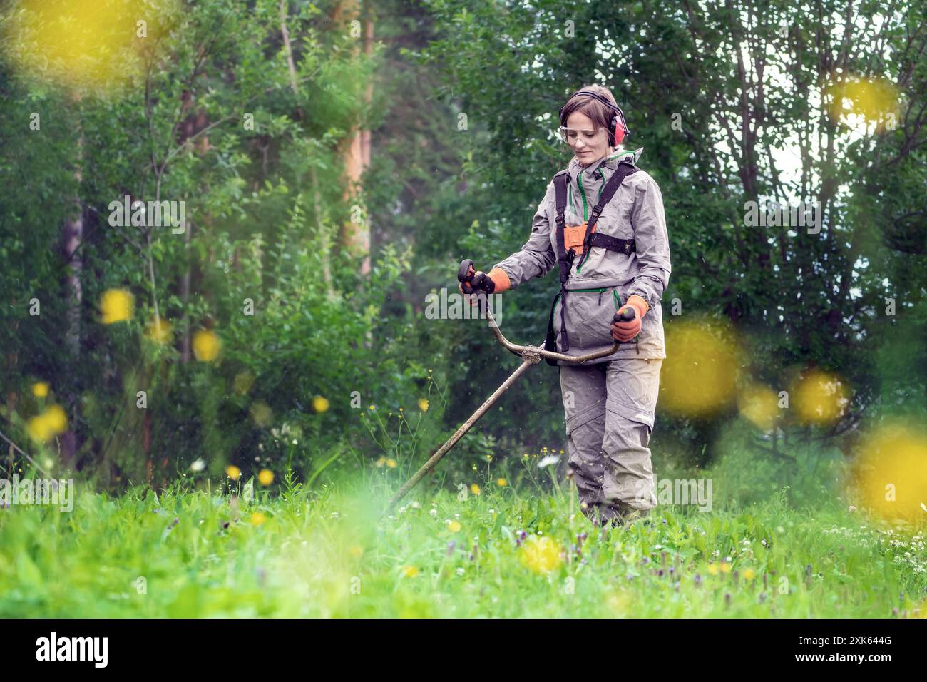 A woman using a brush cutter in a garden with blooming plants, doing ...