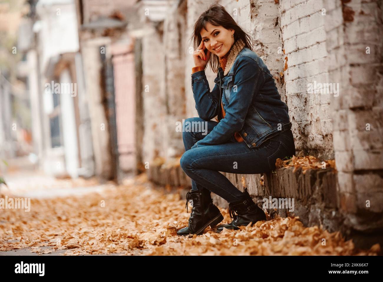 A young woman wearing a denim jacket and jeans sits on a ledge against ...