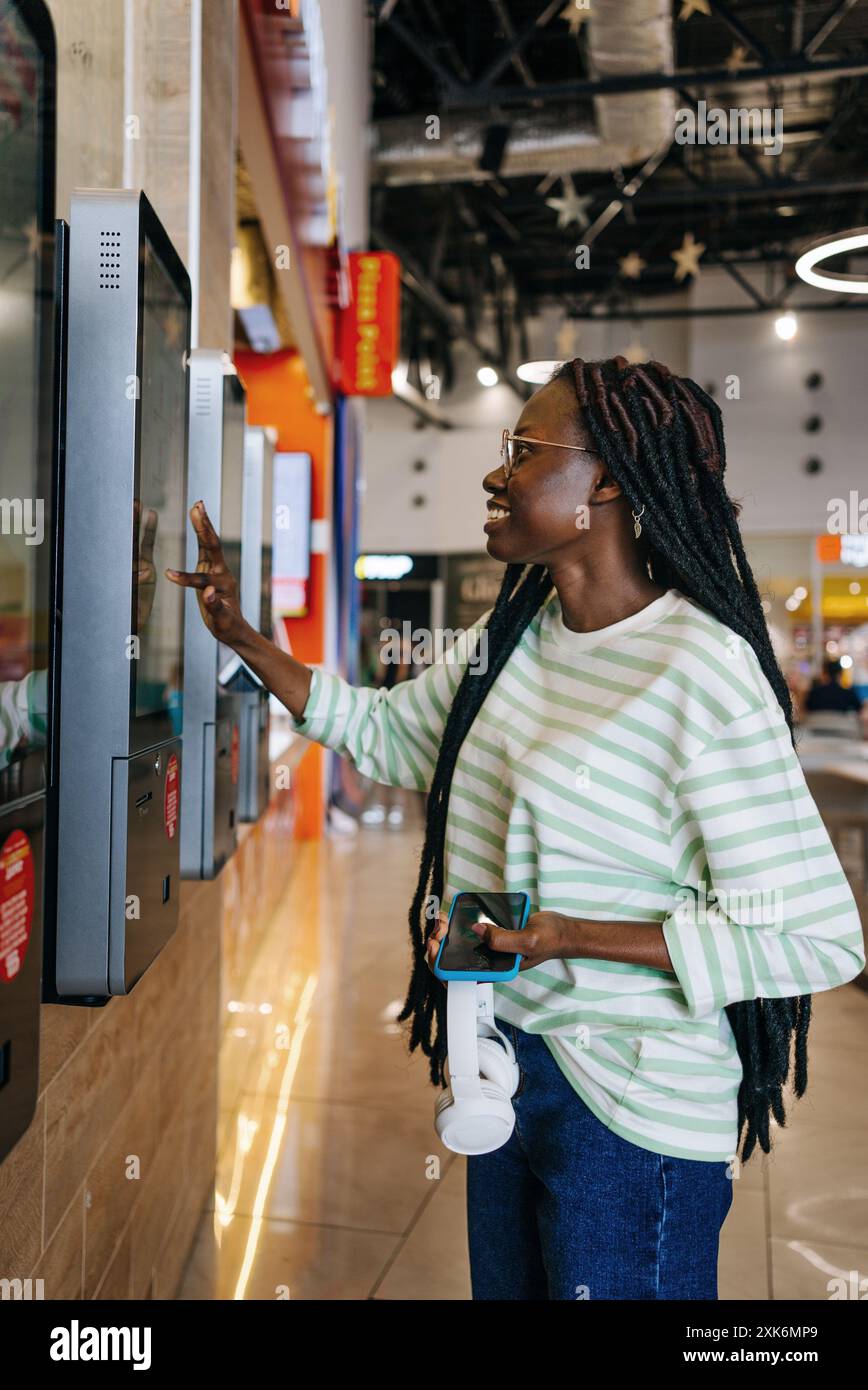 African American girl Using Self-Service Kiosk at Fast Food Restaurant. Black Woman in striped ...