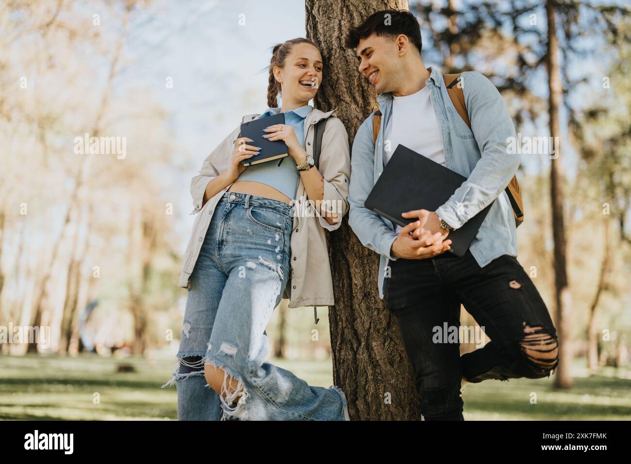 Happy students leaning against a tree with books in a park setting ...