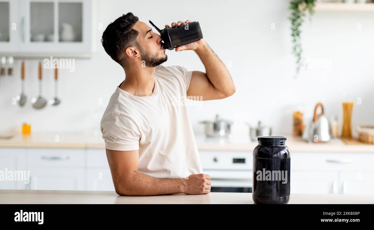 Young Arab guy drinking protein shake from bottle at kitchen, copy ...