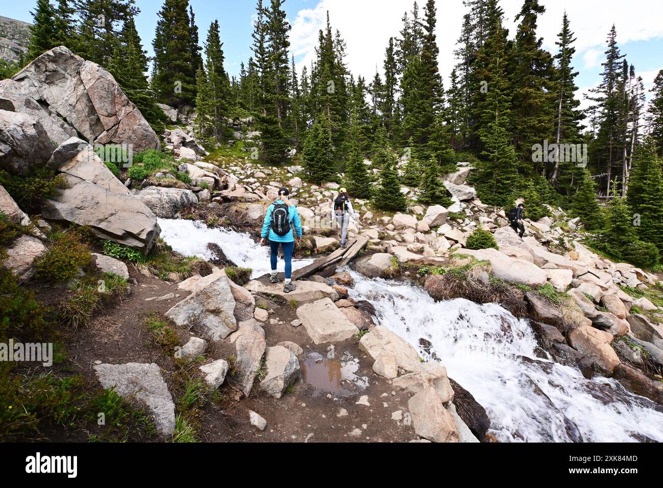 Three hikers crossing stream on narrow log bridge on Lake Isabelle ...